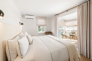 Quarto de hotel com uma cama grande, janela com cortinas, pequena mesa com cadeiras e ar condicionado na parede.