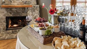 Mesa de buffet com diversos alimentos, copos de vinho e garrafas, com uma lareira de pedra e grandes janelas ao fundo.