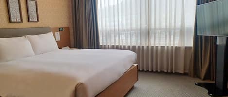 Quarto de hotel com uma cama grande, uma TV de tela plana, uma mesa de cabeceira e vista da cidade através de cortinas finas.