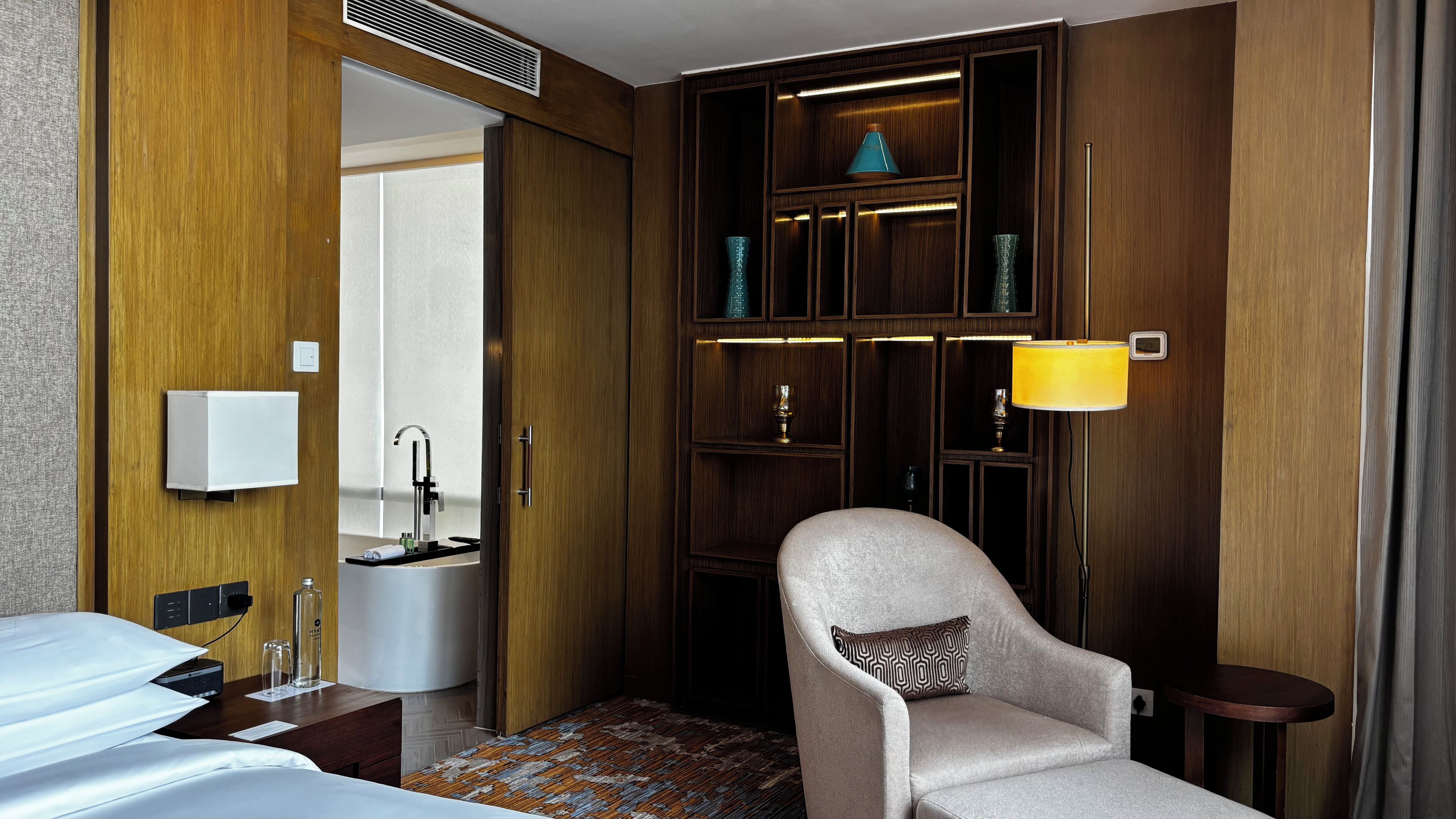 Quarto de hotel com cama, cadeira, escrivaninha, banheiro e prateleira com objetos decorativos.