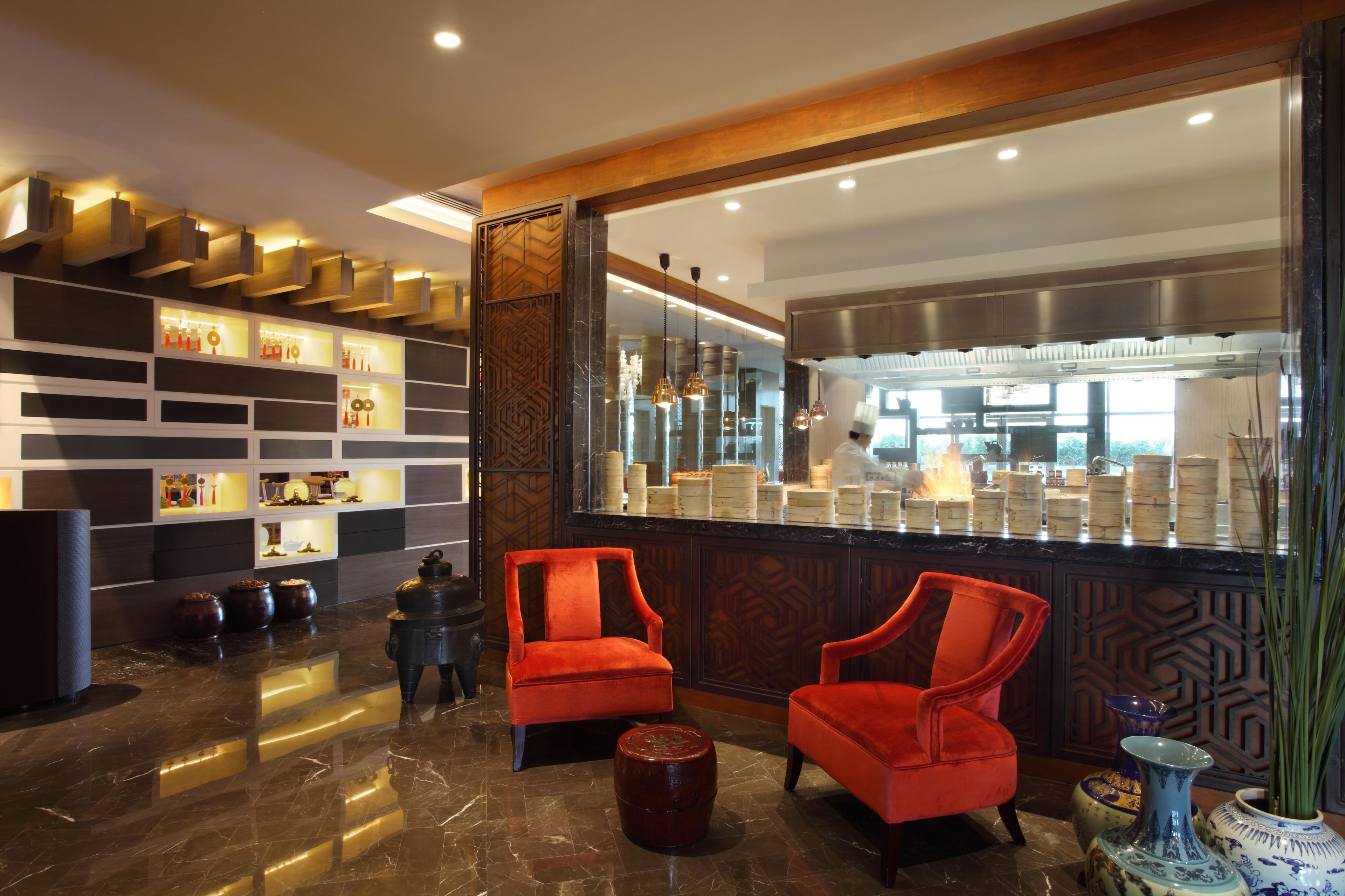 Um lobby com duas poltronas vermelhas, piso de mármore e área de bar com um chef preparando comida.