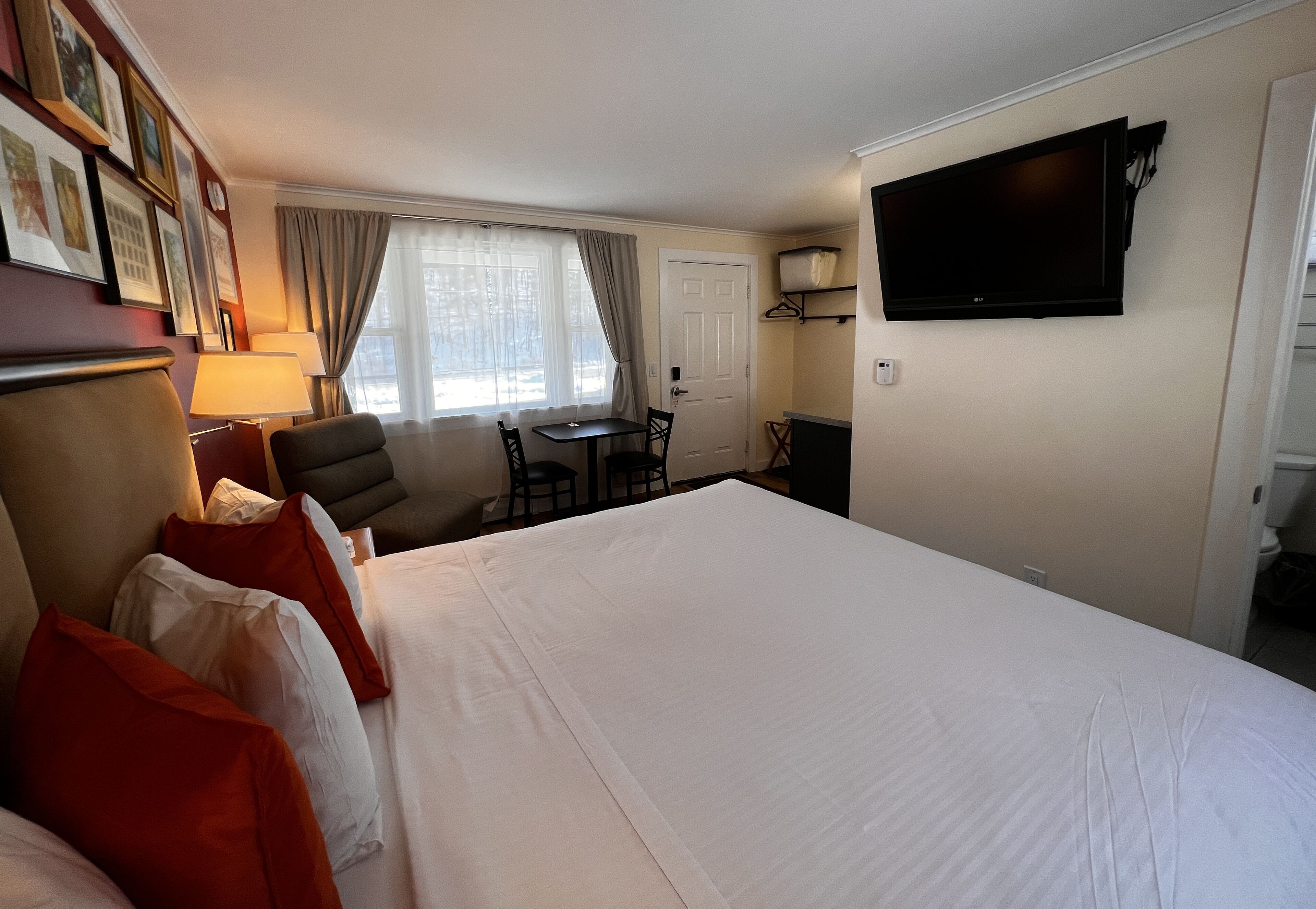 Quarto de hotel com uma cama grande, uma escrivaninha com duas cadeiras, uma televisão fixada na parede e quadros enquadrados.