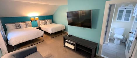 Quarto de hotel com duas camas, televisão, escrivaninha e casa de banho visível através de uma porta aberta.