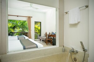 Casa de banho de hotel com um espelho grande, uma banheira, um chuveiro e vista para uma varanda com uma cama e uma secretária.
