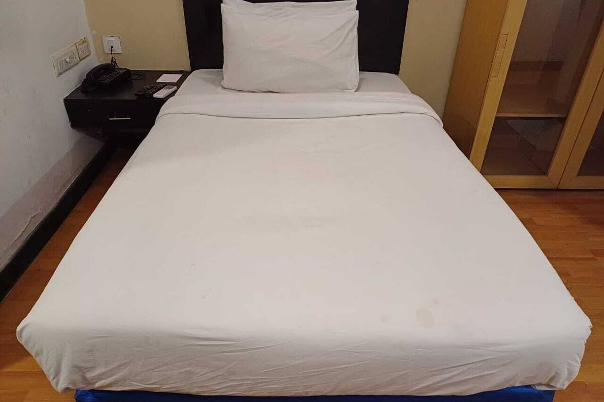 Uma cama de solteiro com colchão e travesseiro brancos, uma mesa de cabeceira com telefone e papéis, e piso de madeira.