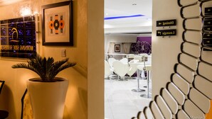 Um hall de hotel moderno com uma grande planta em vaso, obras de arte emolduradas e placas indicativas para os espaços de refeições e lounge.