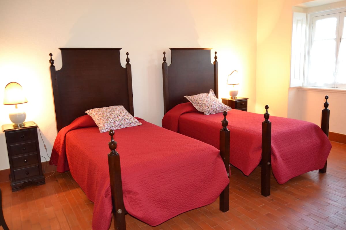 Quarto com duas camas de madeira, cada uma com colcha e travesseiro vermelhos. Há mesas de cabeceira com abajures e uma janela com persianas brancas.
