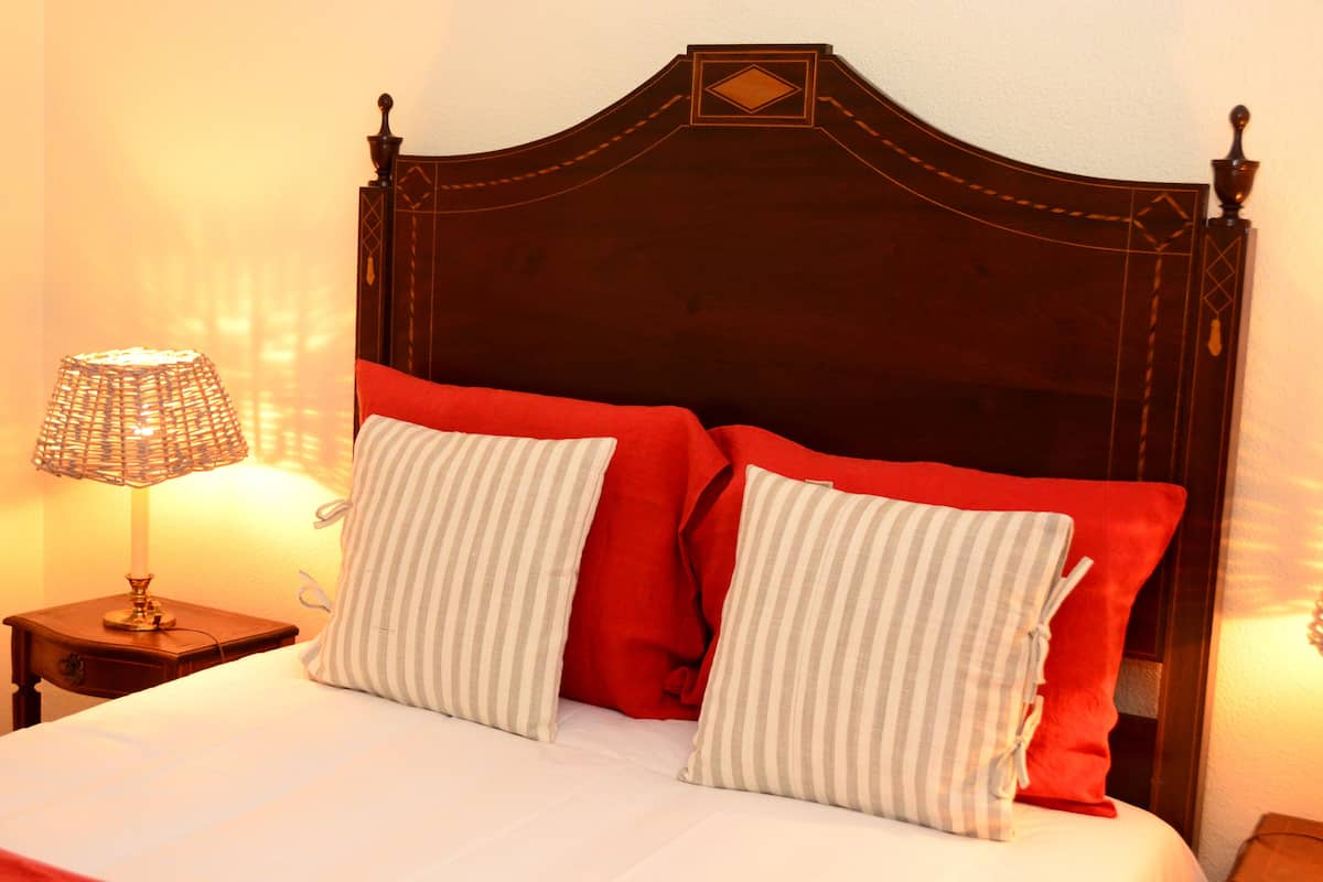 Cama bem arrumada com almofadas listradas em vermelho e branco, cabeceira de madeira e mesa de cabeceira com luminária.