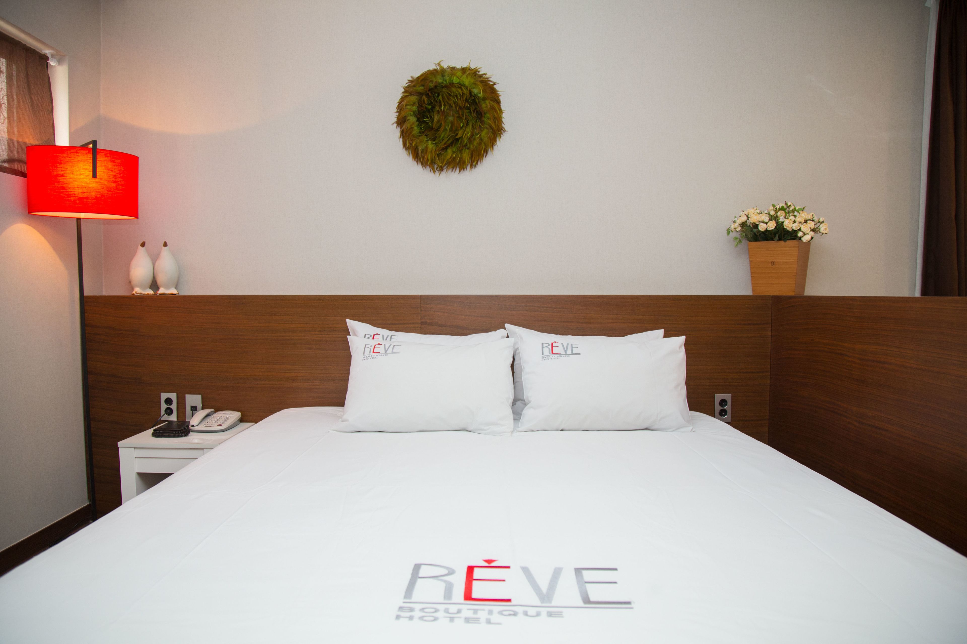 Quarto de hotel com cama, mesa de cabeceira com telefone, abajur vermelho e uma coroa pendurada na parede.