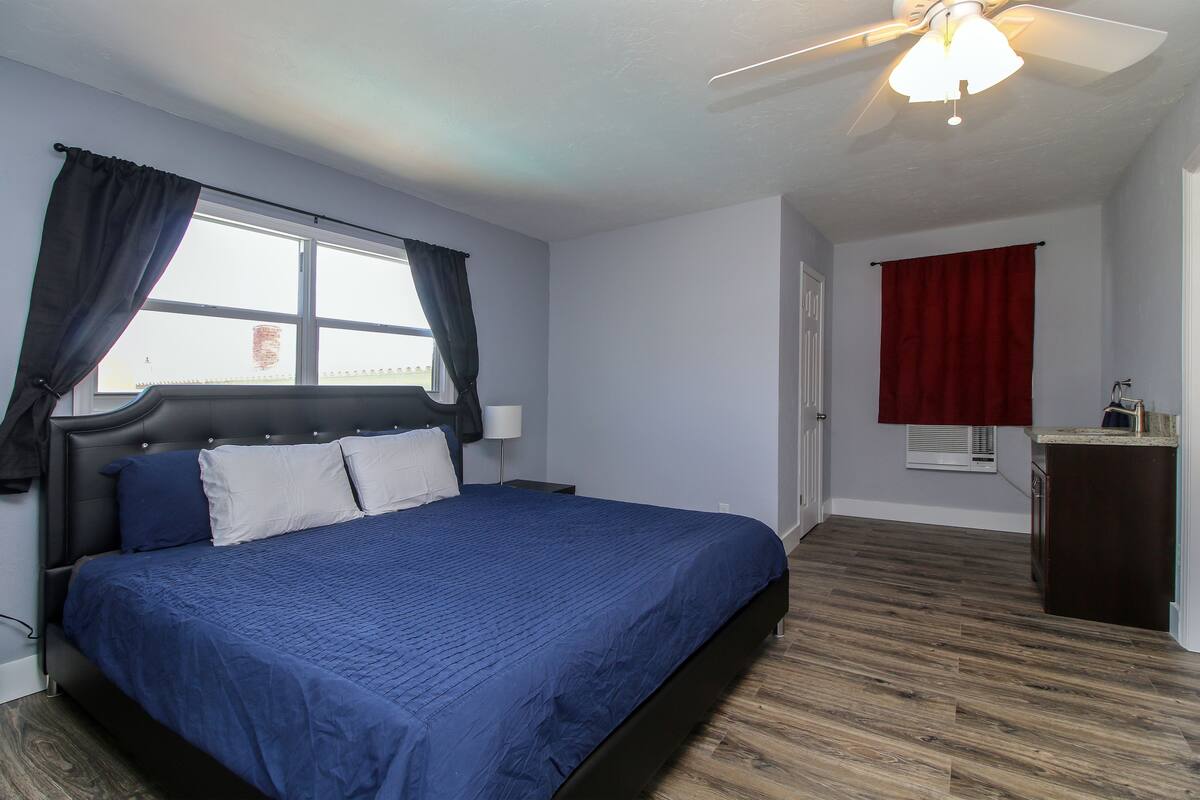 Quarto com cama, ventoinha no teto, janela com cortinas e vista para um edifício.