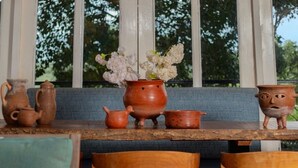 Uma mesa de madeira com vários vasos de barro e um vaso com flores, em frente a um sofá azul e uma janela com vista para árvores.