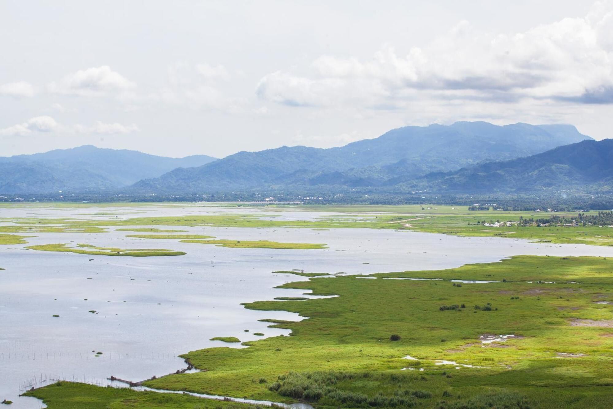 Paisagem com rio, campos verdes e montanhas ao fundo.