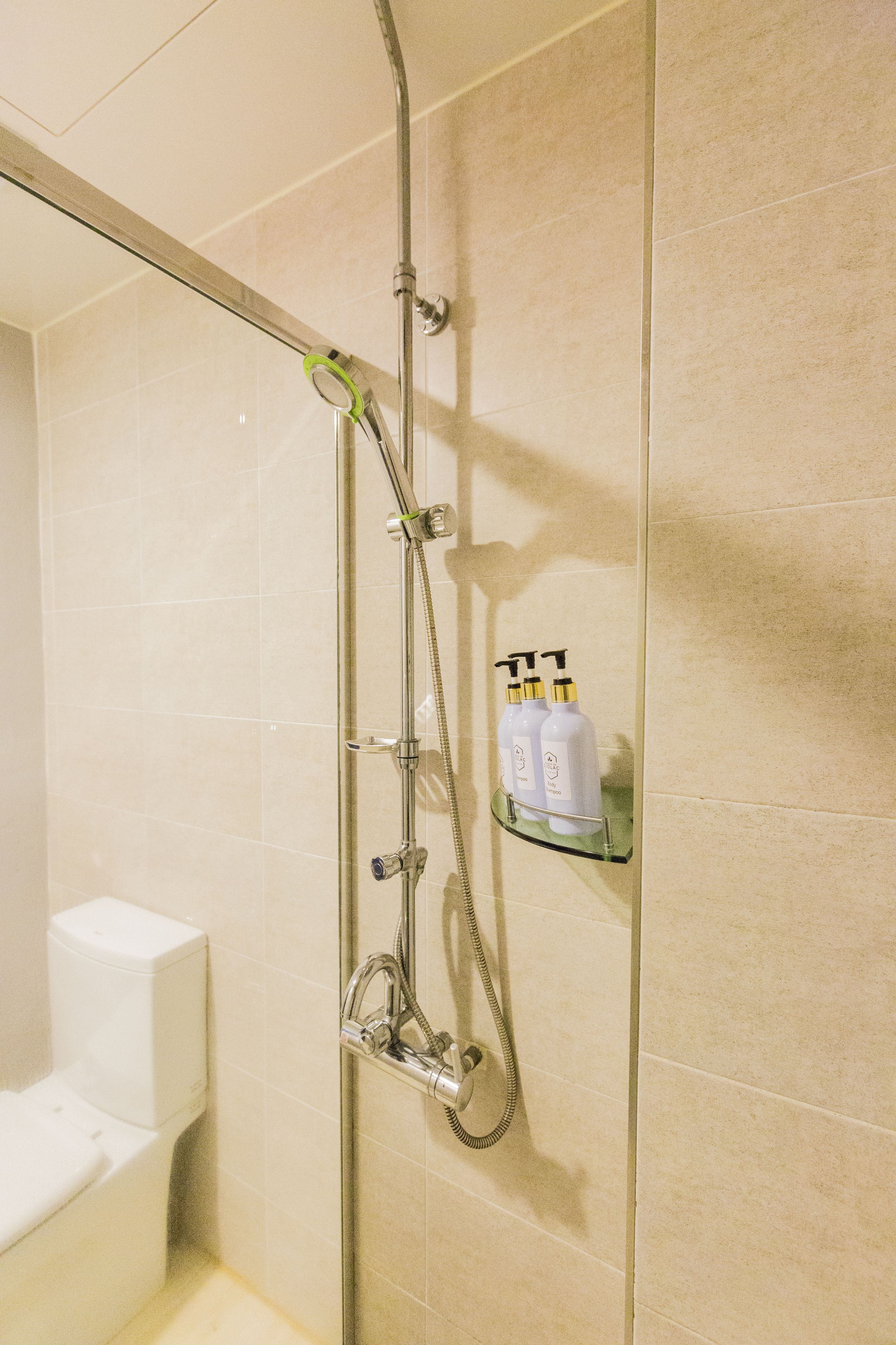Chuveiro com chuveiro manual e torneira fixada na parede. Há uma prateleira com duas garrafas de shampoo e condicionador.