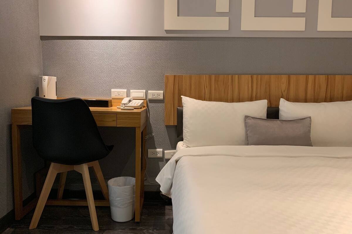 Quarto de hotel com escrivaninha de madeira, uma cadeira, telefone e cama com almofadas.