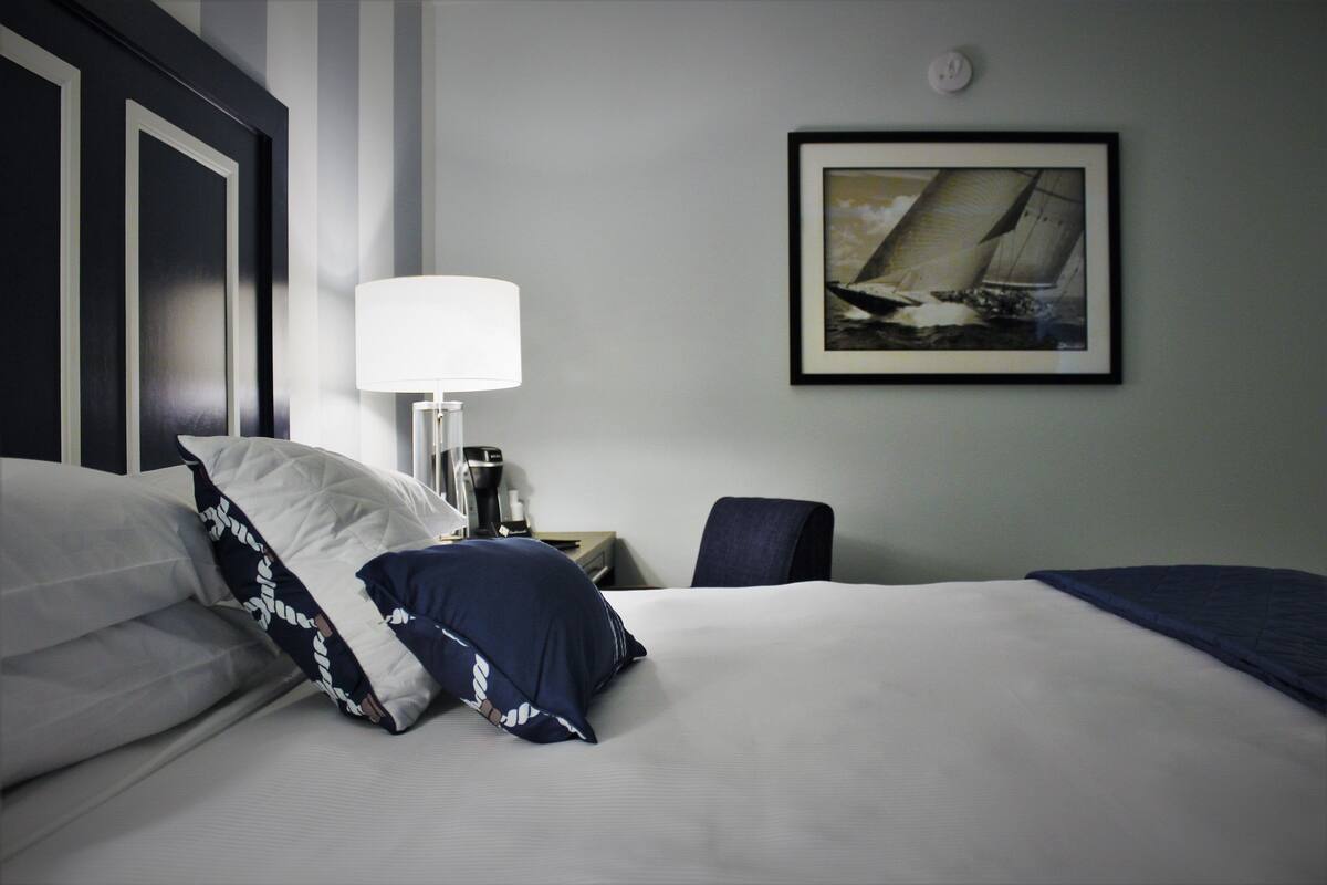 Quarto de hotel com uma cama bem arrumada, mesa de cabeceira com abajur, quadro na parede e uma cadeira.