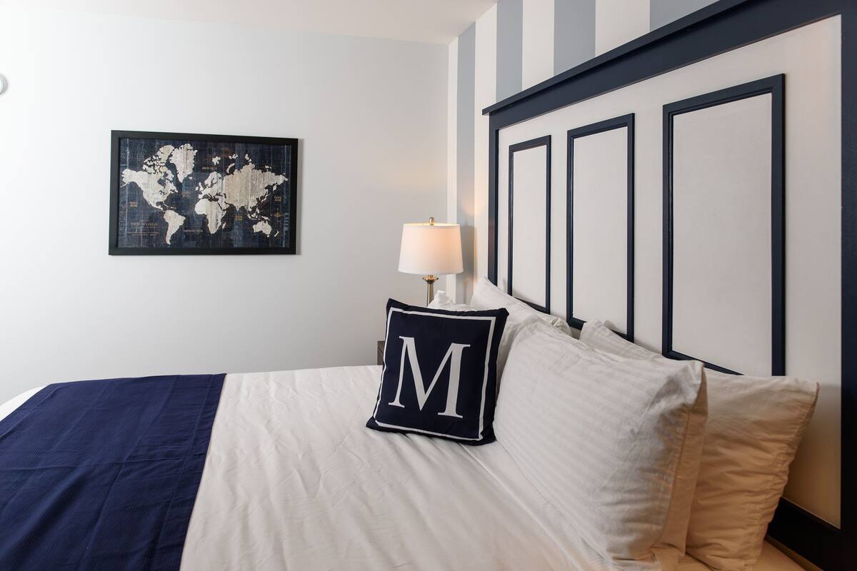Cama bem arrumada com colcha azul marinho e branca, travesseiro com uma grande letra 'M' e um mapa-múndi na parede.