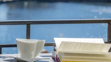 Uma mesa com uma xícara branca e um livro aberto, com vista para um corpo de água e prédios ao fundo.