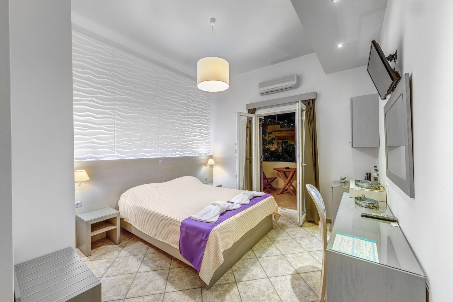 Quarto com cama, mesas de cabeceira, escrivaninha, cadeira, janela com persianas, ar condicionado e varanda com mesa e cadeiras.