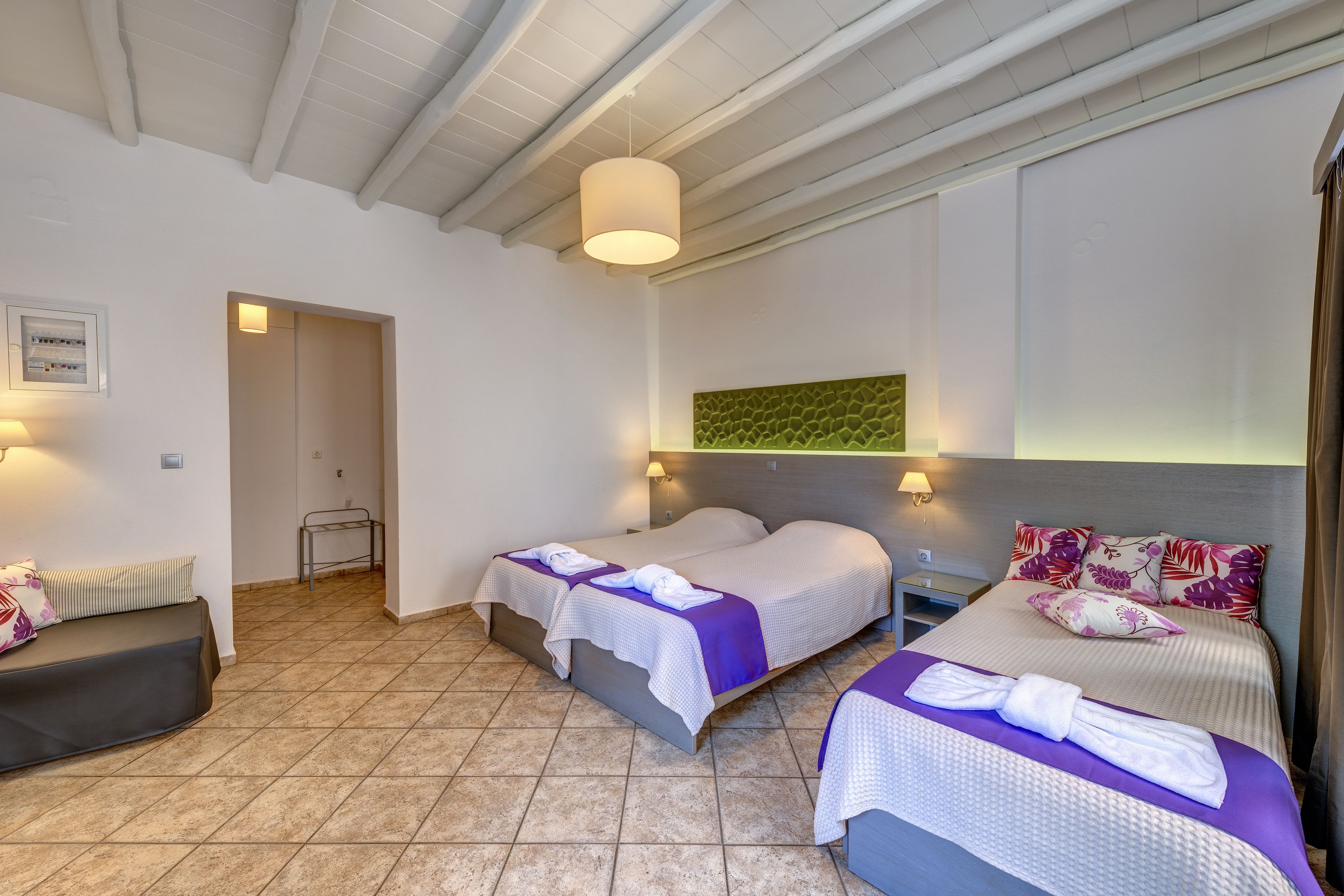 Quarto de hotel com duas camas, um sofá, uma mesinha e um candeeiro fixado na parede.