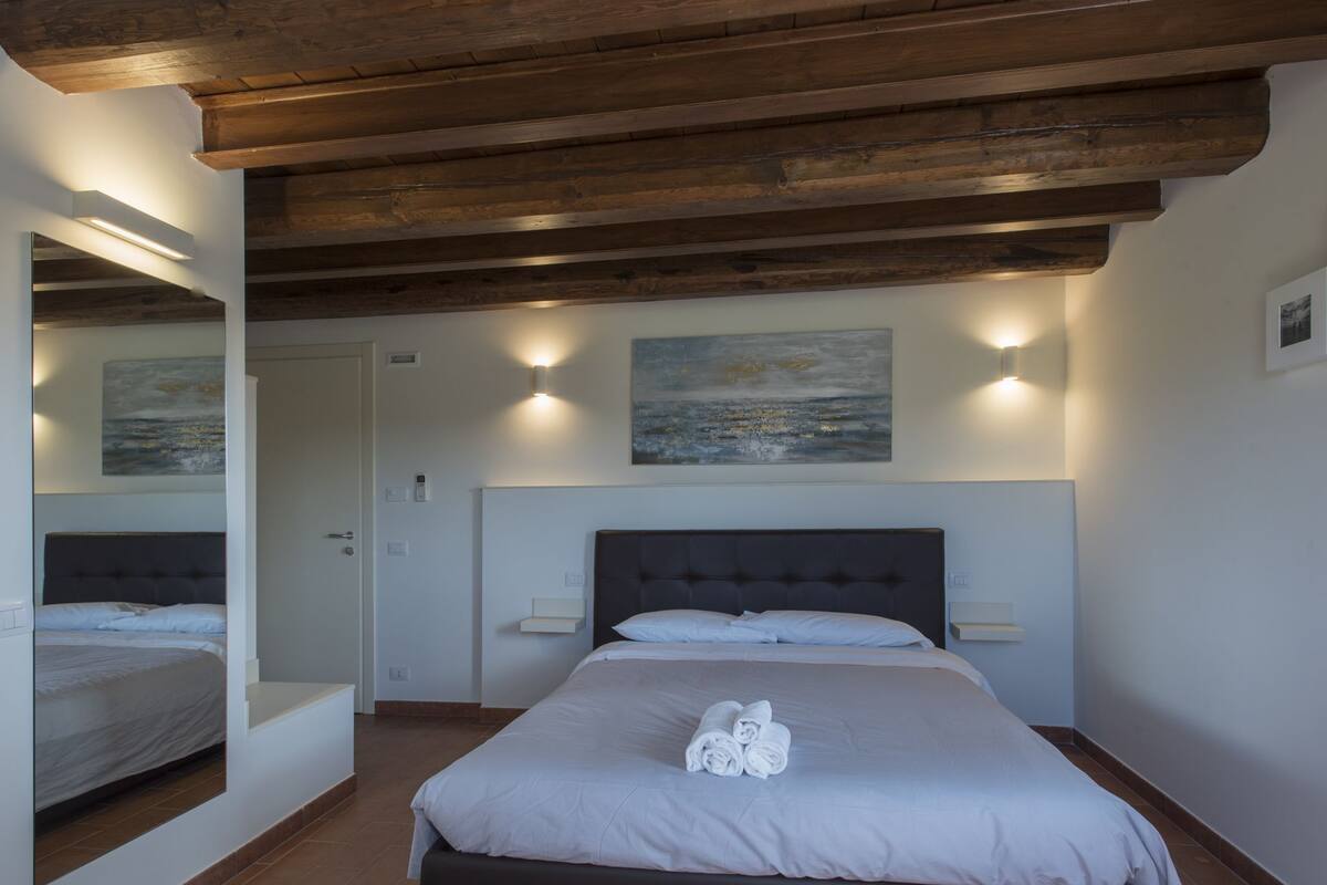 Quarto com cama grande, vigas de madeira no teto, pintura na parede e espelho à esquerda.