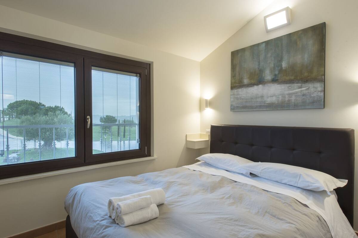 Quarto com cama, duas janelas, um quadro e uma mesinha de cabeceira.