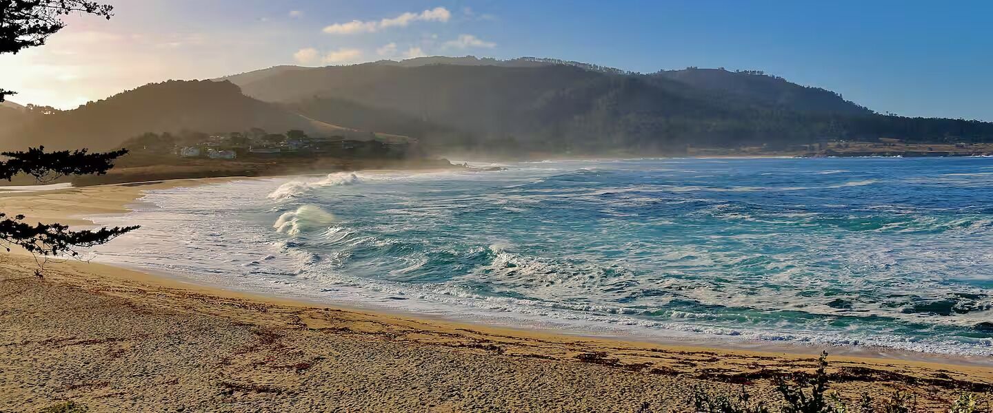 Uma praia com ondas, céu claro e paisagem montanhosa ao fundo.