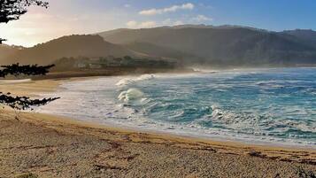 Uma praia com ondas, céu claro e paisagem montanhosa ao fundo.