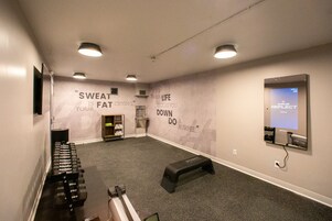 Uma academia com racks de pesos, bebedouro, frases motivacionais na parede e uma tela digital exibindo um programa de exercícios.