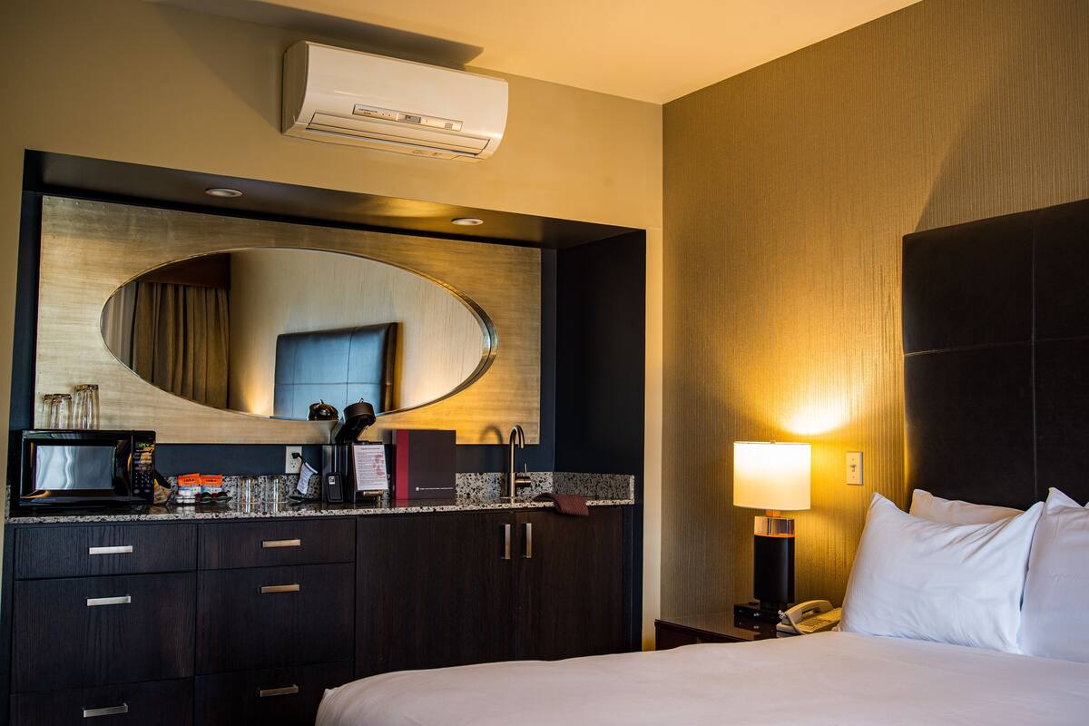 Quarto de hotel com cama, um espelho grande, micro-ondas, pia e mesa de cabeceira com abajur.