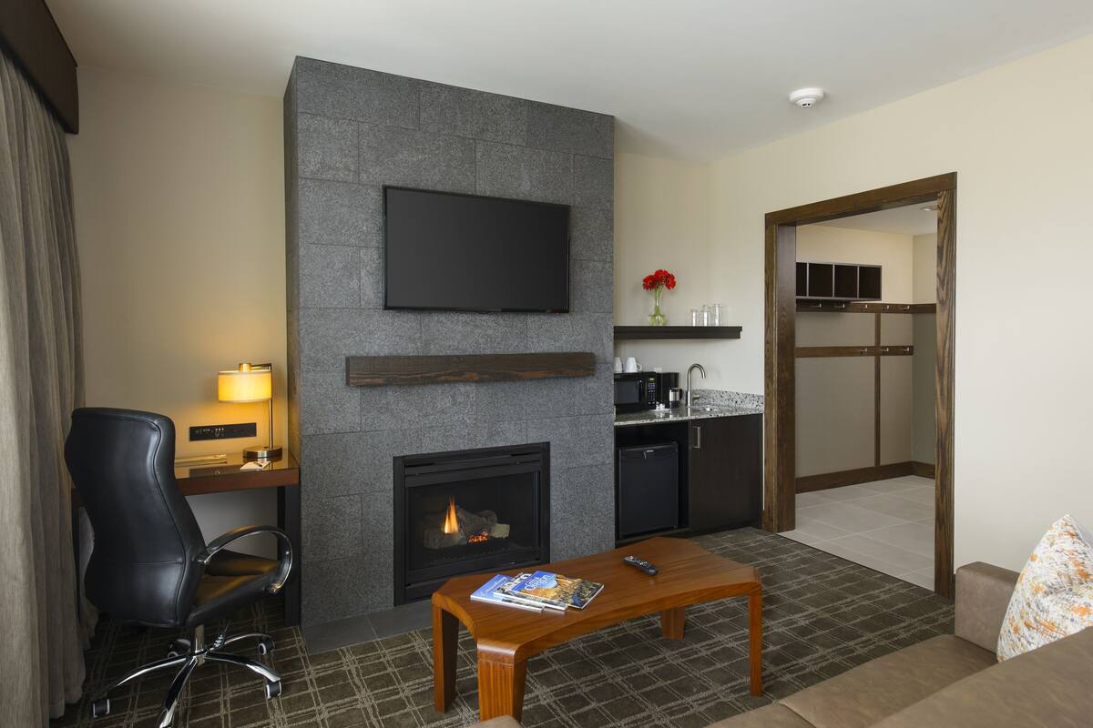 Quarto de hotel com televisão de tela plana fixada em uma parede de pedra, lareira, escrivaninha com luminária, máquina de café e sofá.