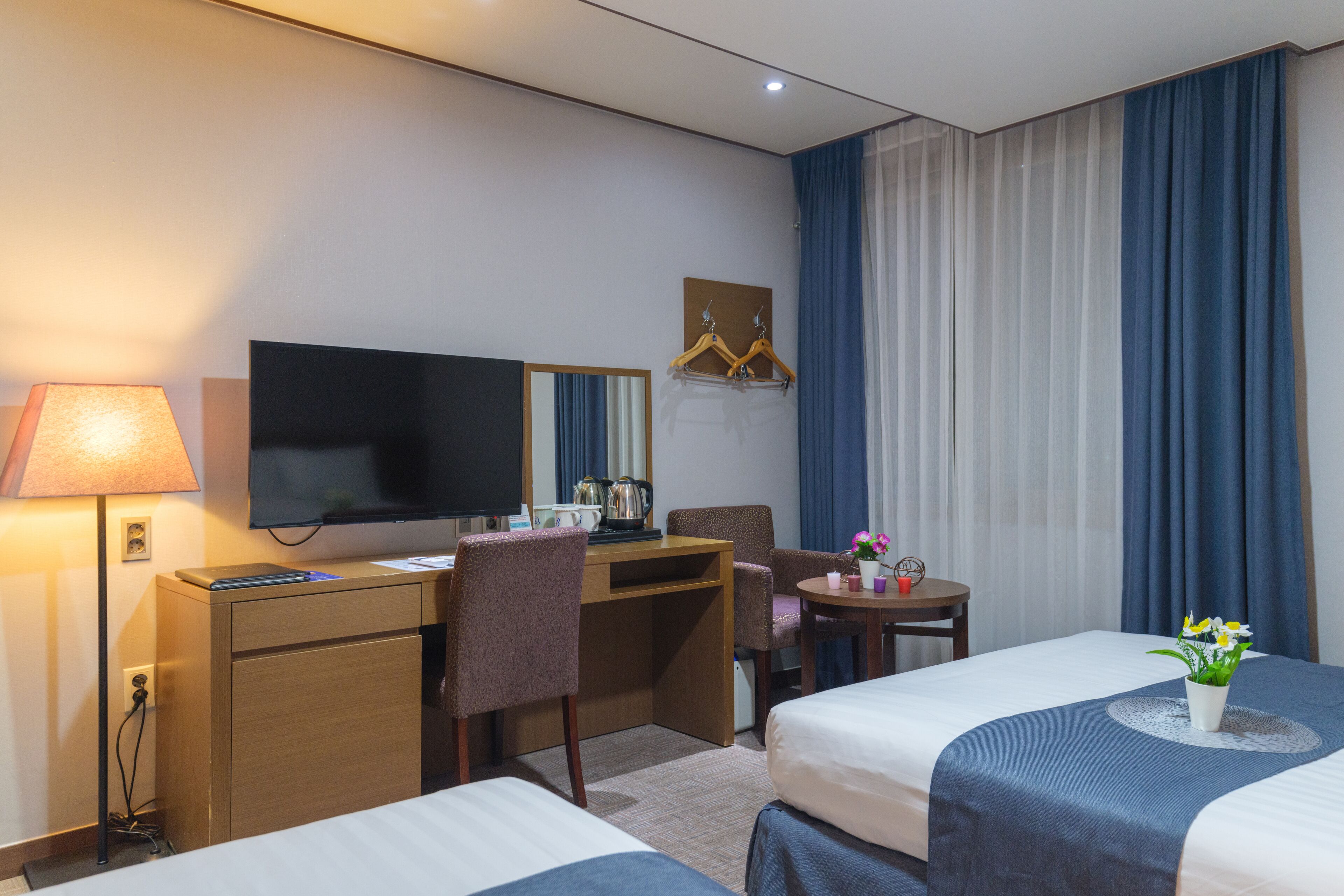 Quarto de hotel com duas camas, uma escrivaninha, televisão, uma cadeira, uma mesinha de centro com uma chávena e flores, e um abajur.