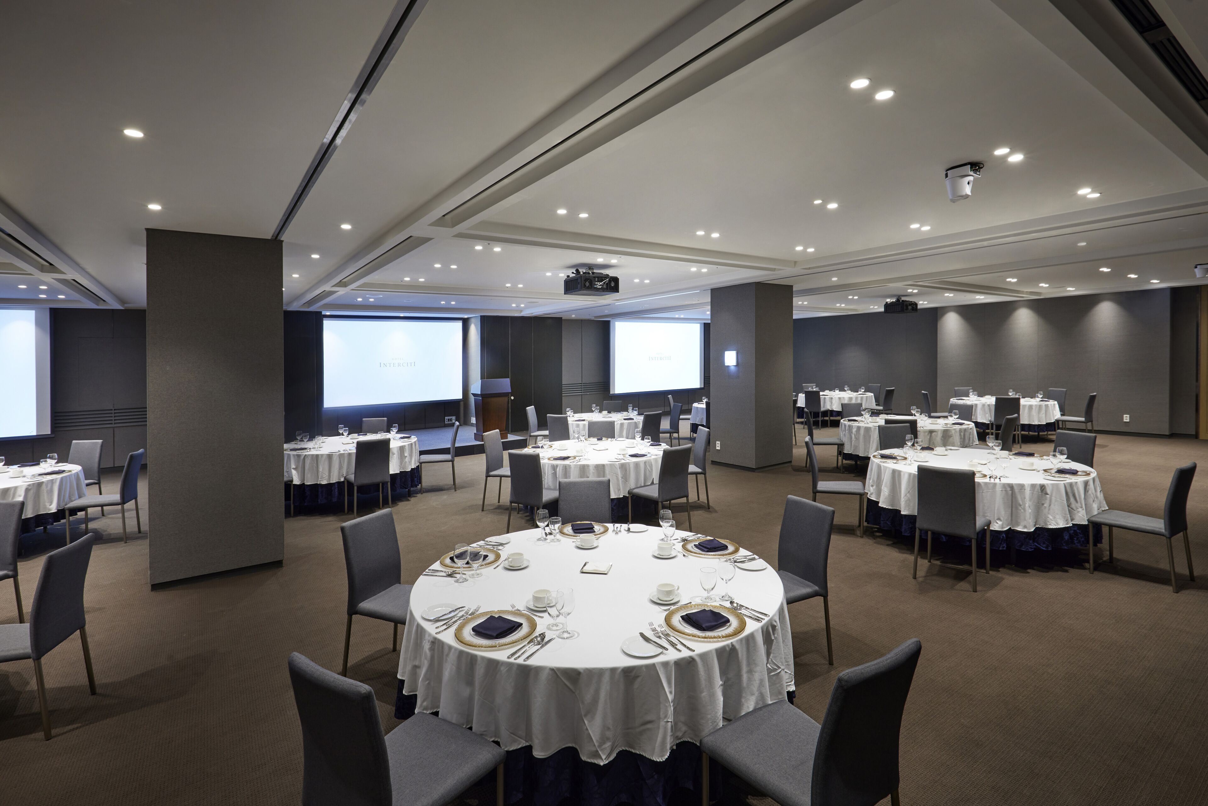 Uma sala de conferências com várias mesas redondas, cada uma com toalhas brancas, pratos dourados e talheres.