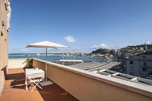 Varanda com mesa e cadeira brancas, um guarda-sol branco e vista para um porto e paisagem urbana.