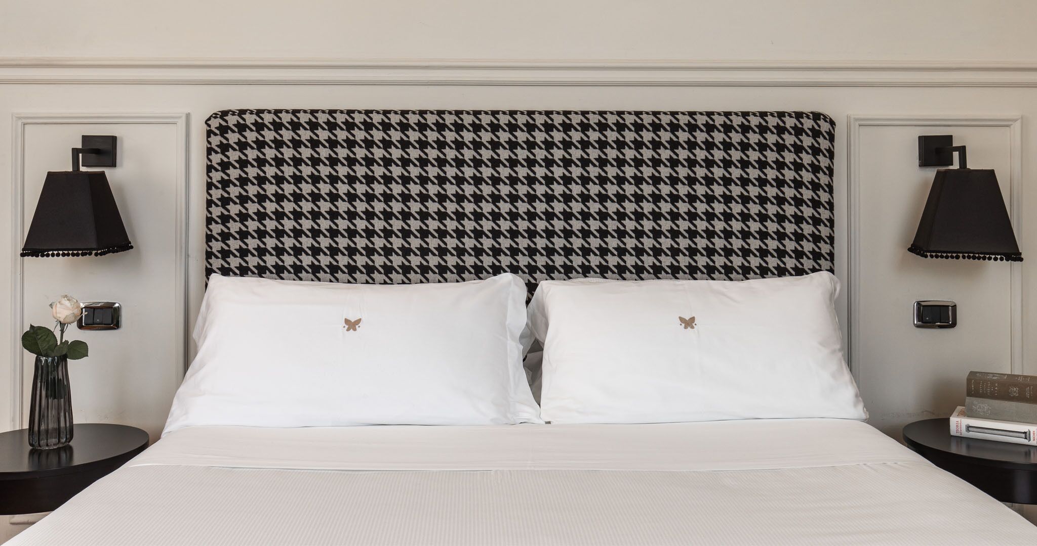 Cama com roupa de cama branca, cabeceira estampada em xadrez houndstooth e duas luminárias de cabeceira.