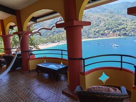 Varanda com vista para uma praia e barcos, com pilares vermelhos e móveis de vime.