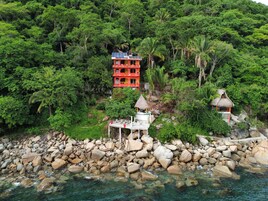 Um edifício vermelho com v árias varandas, rodeado por vegetação exuberante e uma costa rochosa.