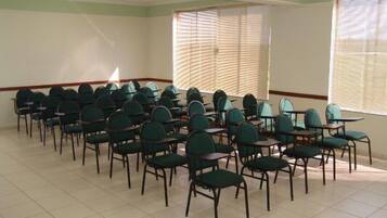 Uma sala de conferências com fileiras de cadeiras e mesas verdes, amplas janelas com persianas e piso revestido de azulejos.