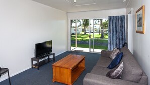 Uma sala de estar moderna com uma televisão de tela plana em um suporte, uma mesa de centro de madeira, um sofá cinza com almofadas e uma grande porta de vidro deslizante que dá para uma área externa gramada.