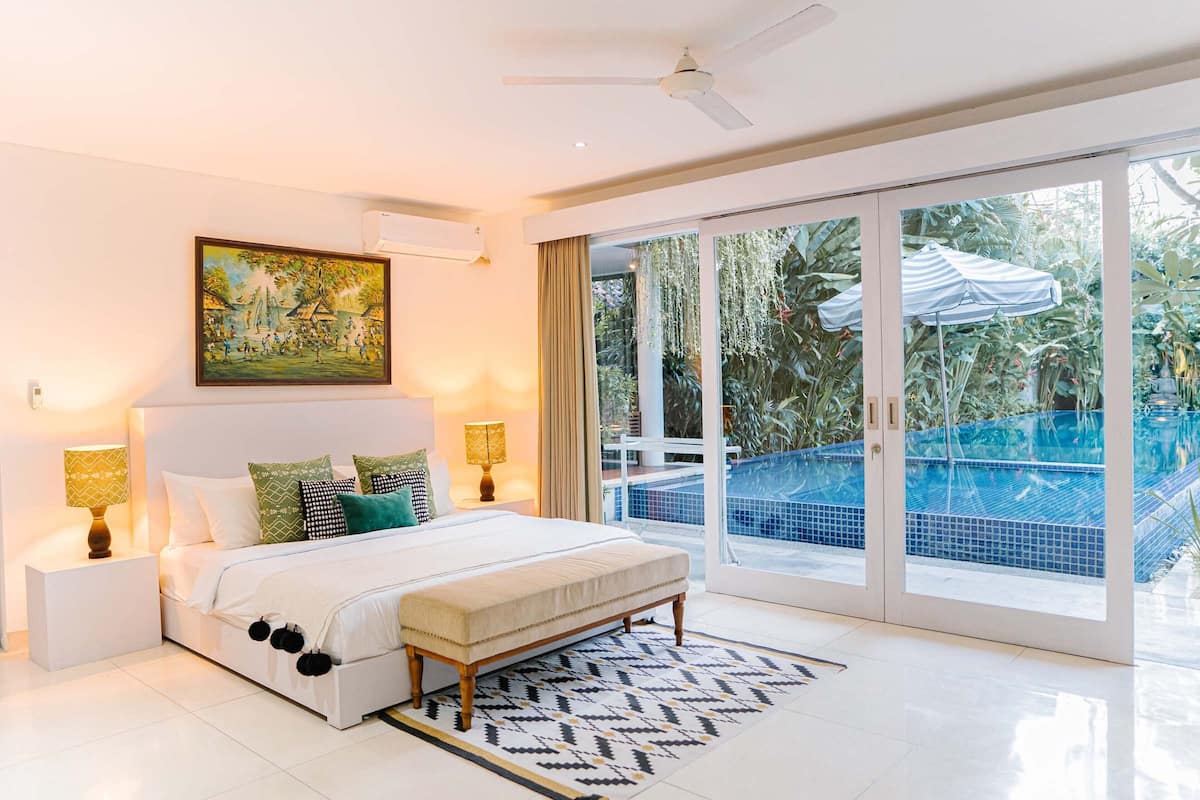 Quarto com uma cama grande, duas luminárias de cabeceira, ventilador de teto e vista para uma piscina através de grandes portas de correr.
