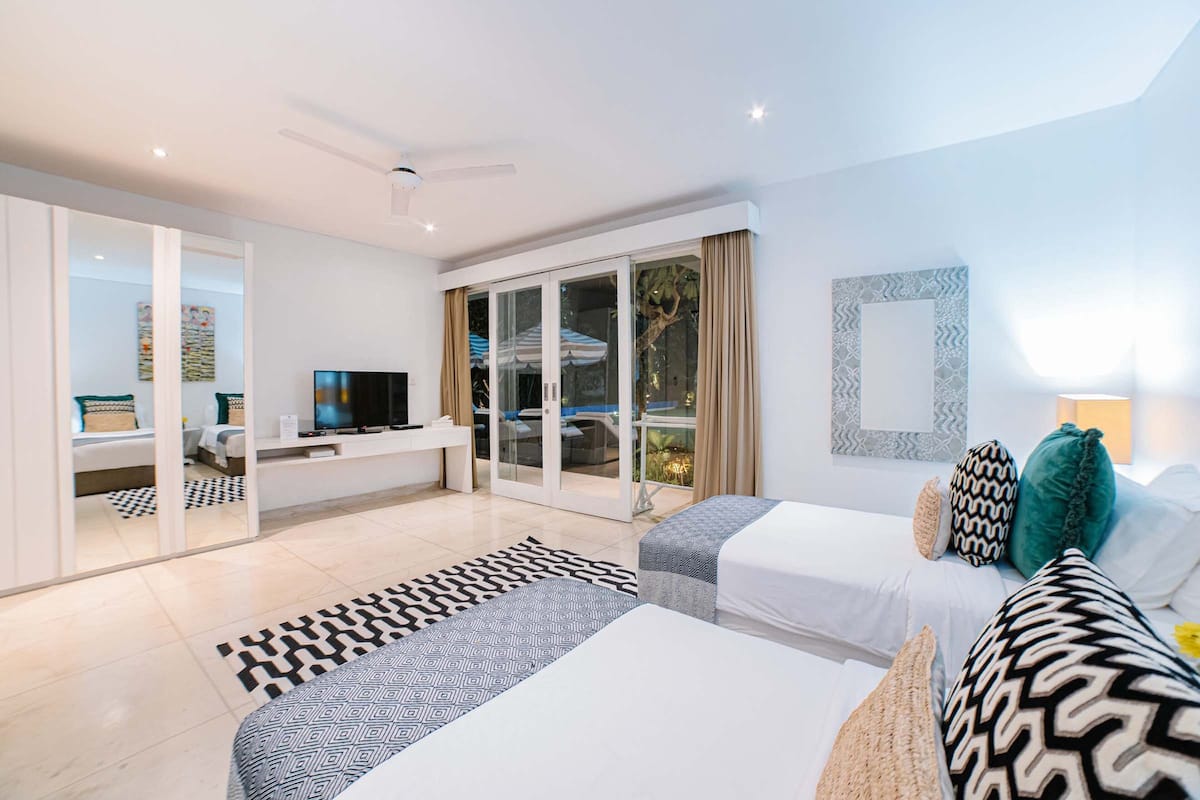 Quarto de hotel moderno com duas camas, uma televisão, porta de vidro deslizante para uma varanda e ventoinha de teto.