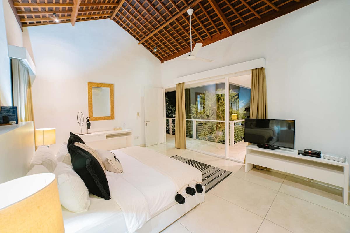 Quarto moderno com uma cama grande, televisão, ventilador de teto e uma porta de vidro deslizante para uma varanda.