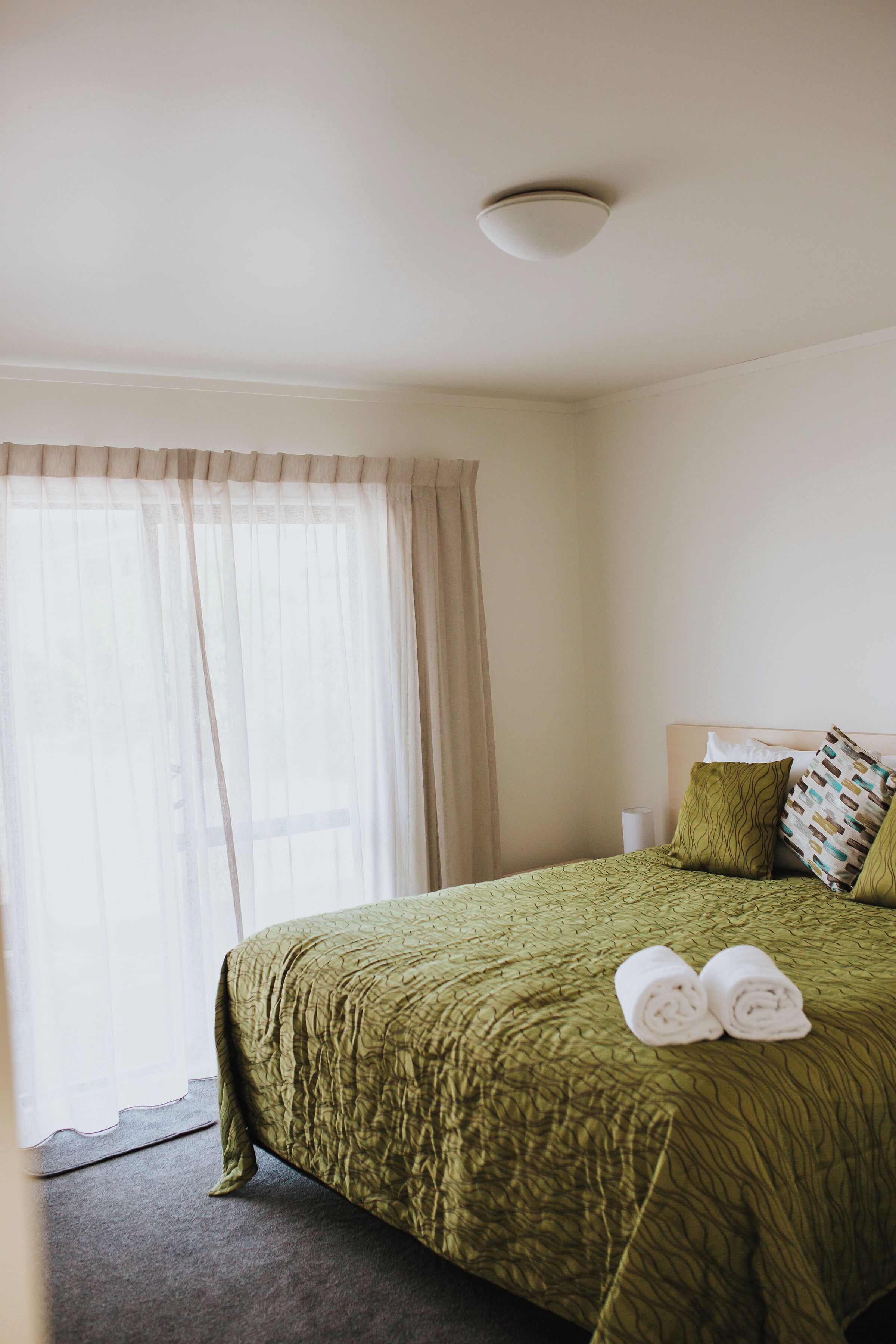 Quarto de hotel com cama, roupa de cama verde, janela com cortinas finas e luminária no teto.