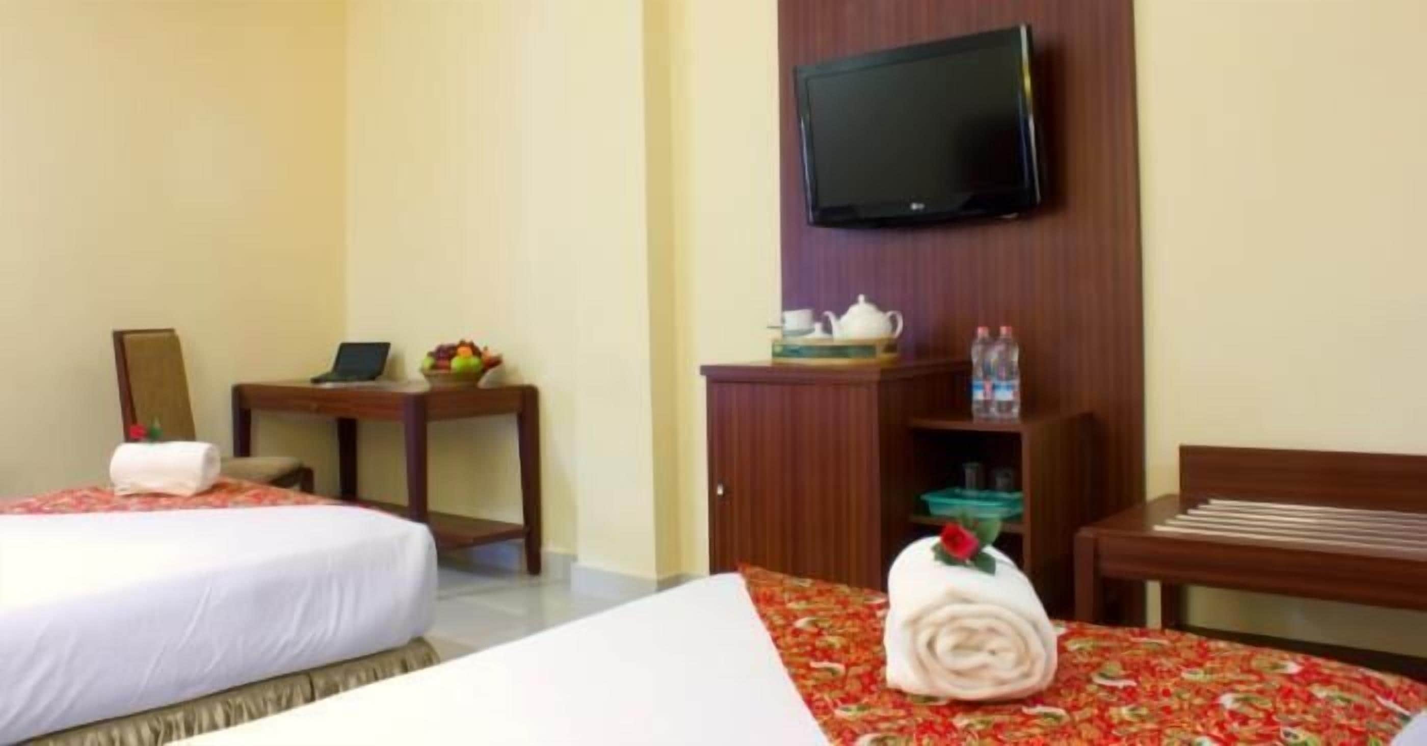 Quarto de hotel com duas camas, uma televisão na parede, uma mesa pequena com um bule e xícaras, e um banquinho.