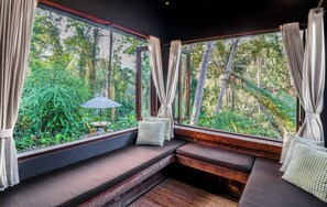 Quarto com janelas amplas, um banco e uma pequena mesa com abajur.