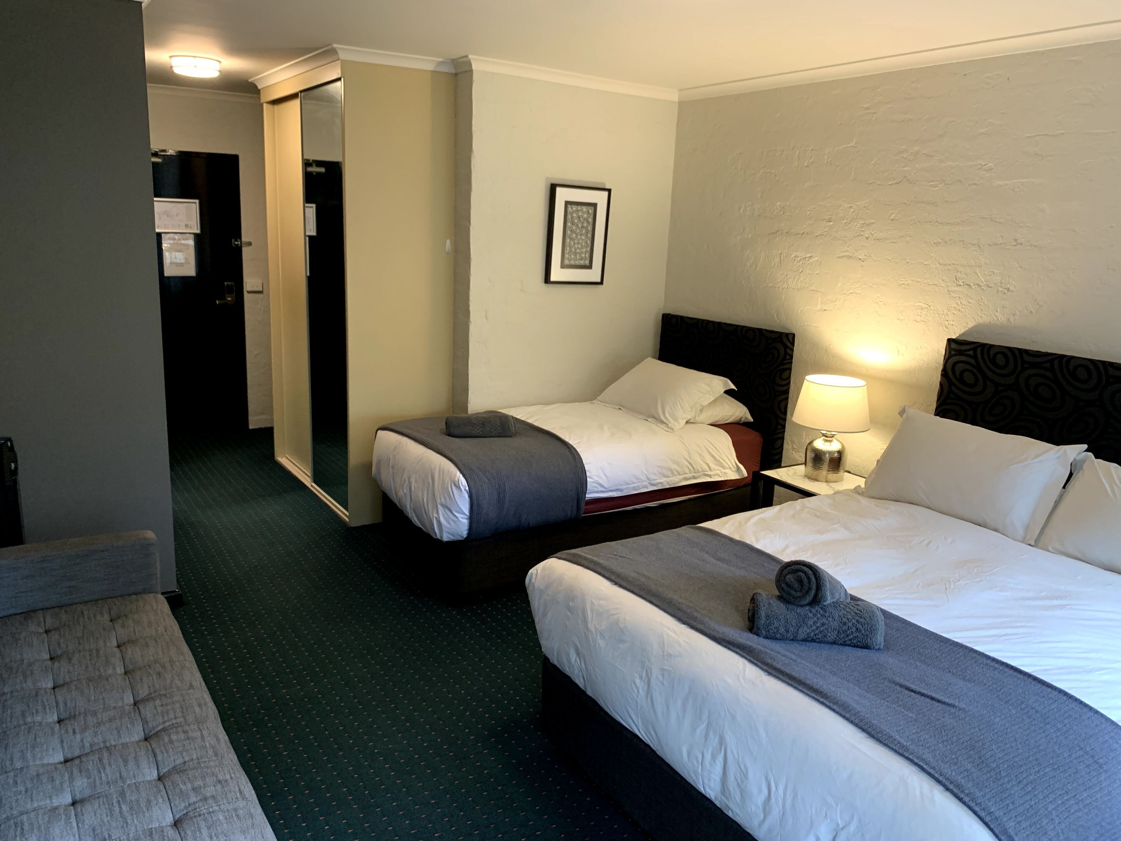 Quarto de hotel com duas camas, uma mesa, uma cadeira e uma porta que dá para um corredor.