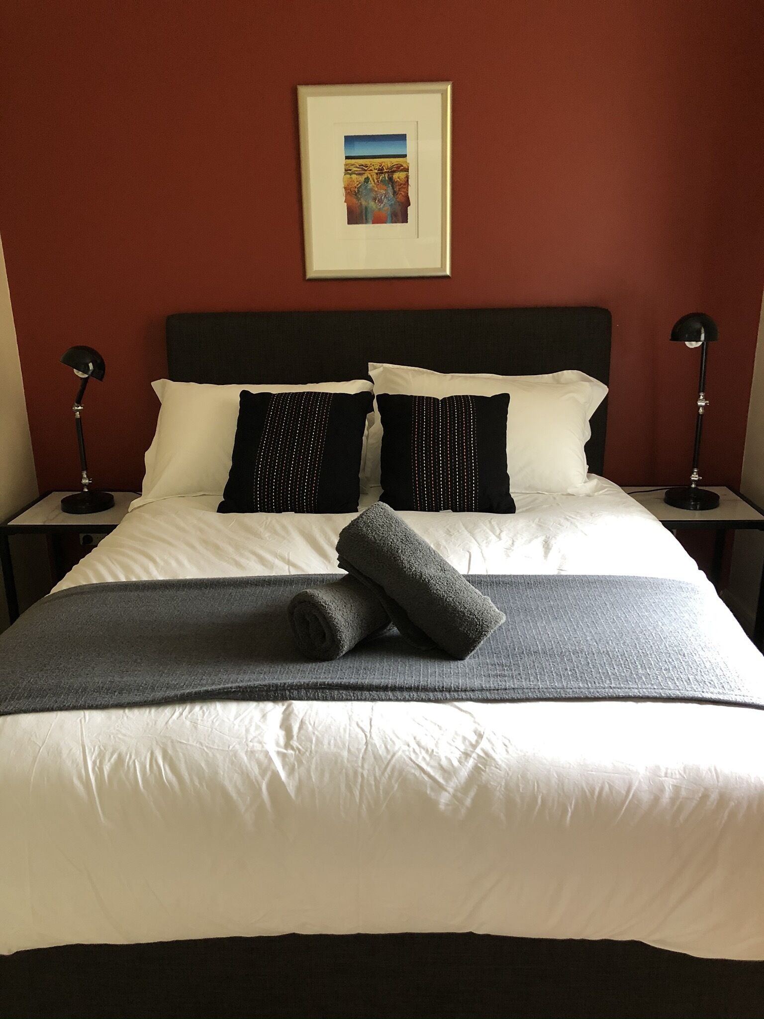 Cama bem arrumada com roupa de cama branca e preta, duas luminárias pretas de cada lado e um quadro na parede.