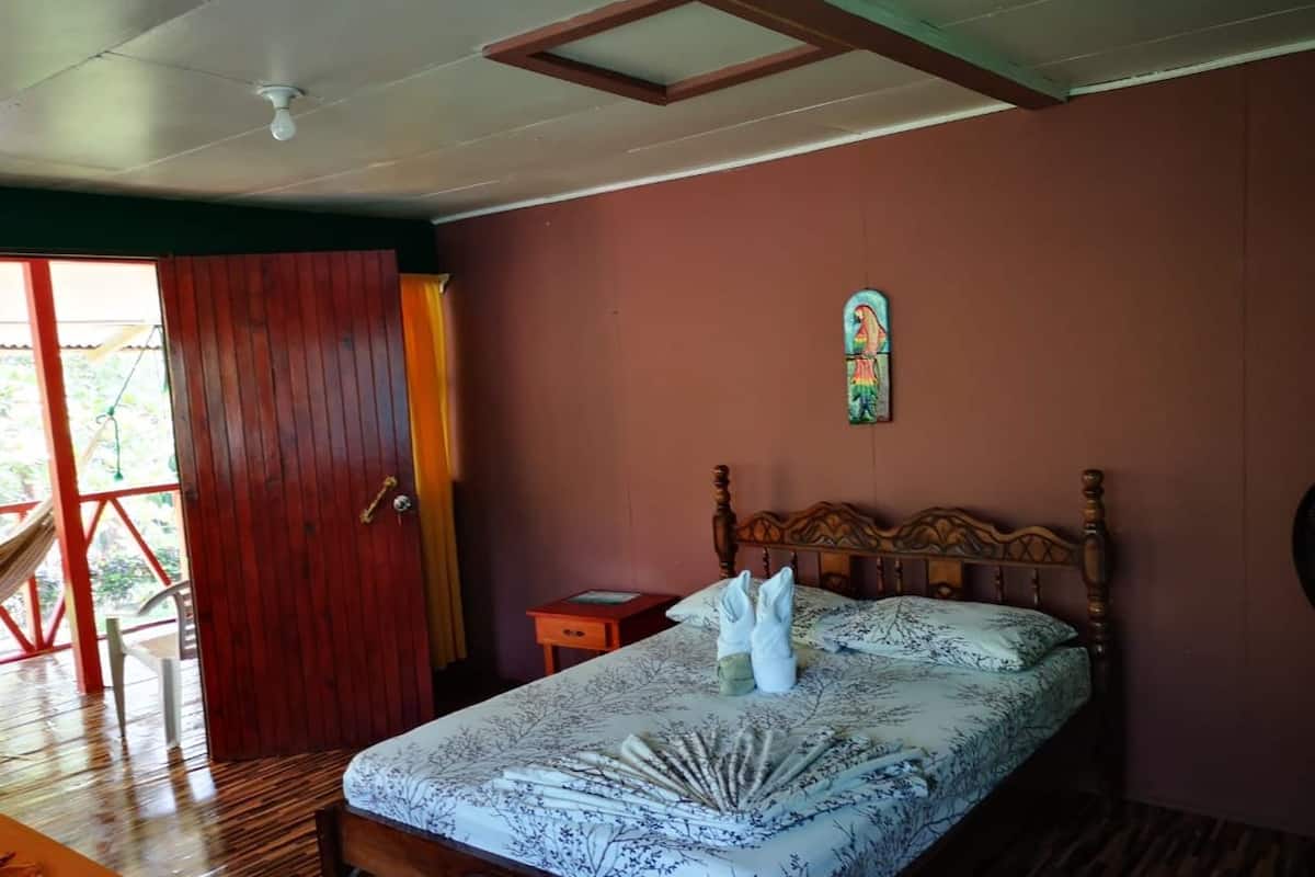 Quarto com cama de madeira, uma pequena mesa de cabeceira e vista para o exterior através de uma porta.
