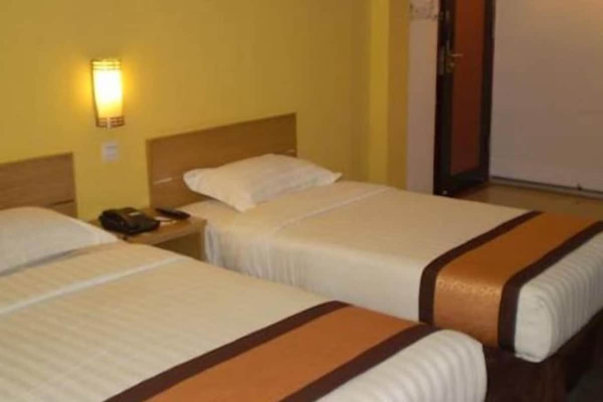Quarto de hotel com duas camas, mesa de cabeceira e uma luminária, além de uma porta.