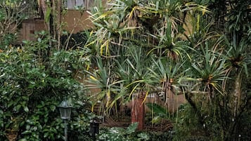 Um jardim com mesa e cadeiras no alpendre, cercado por vegetação exuberante e um edifício tradicional com telhado de azulejos.