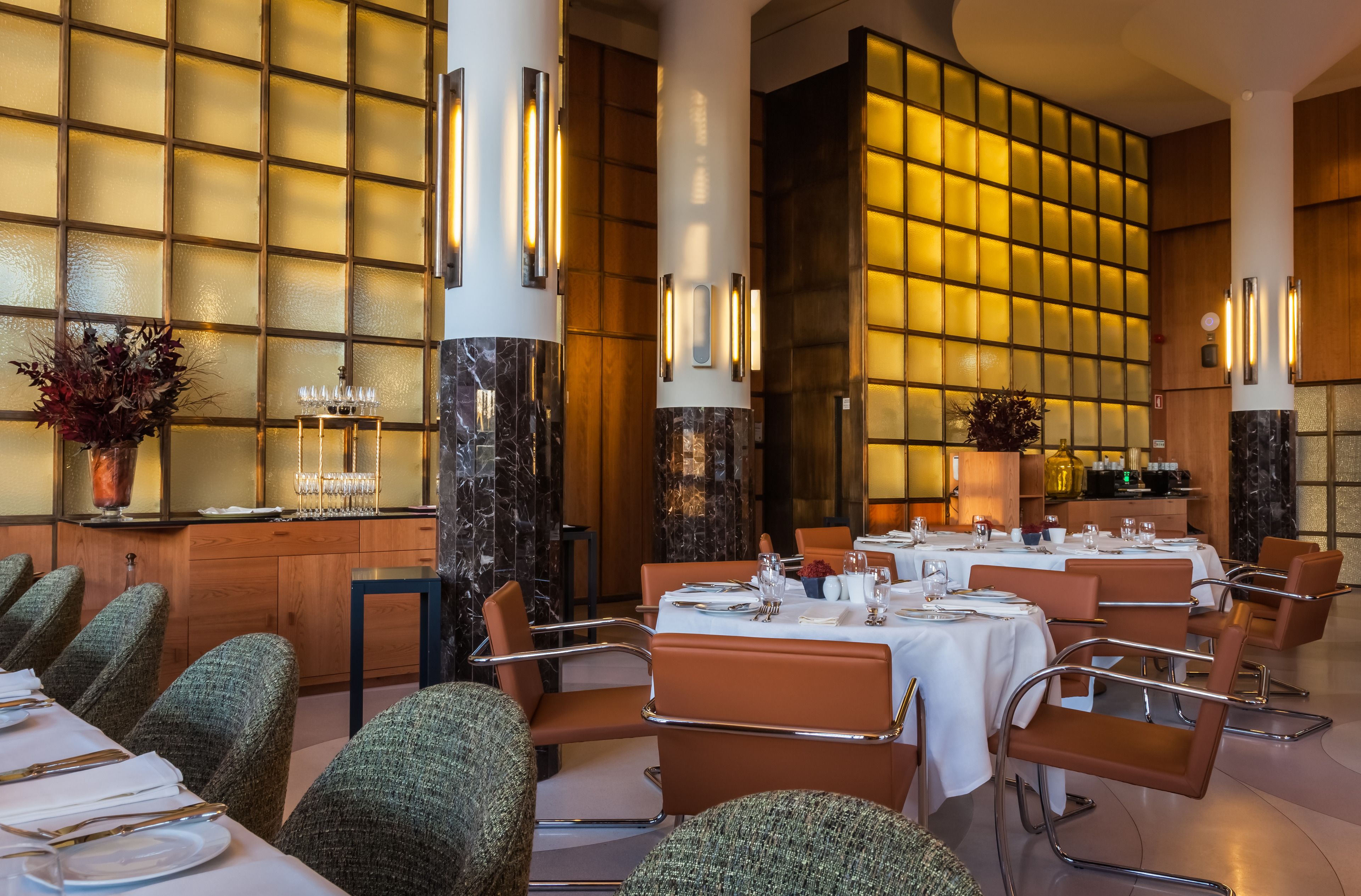 Uma sala de jantar moderna com mesas preparadas para vários convidados, com cadeiras contemporâneas e uma parede decorada em mármore.